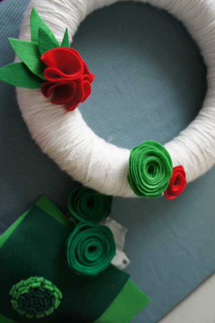 White rope-wrapped wreath with red felt roses and green spiral flowers on a blue textured surface.