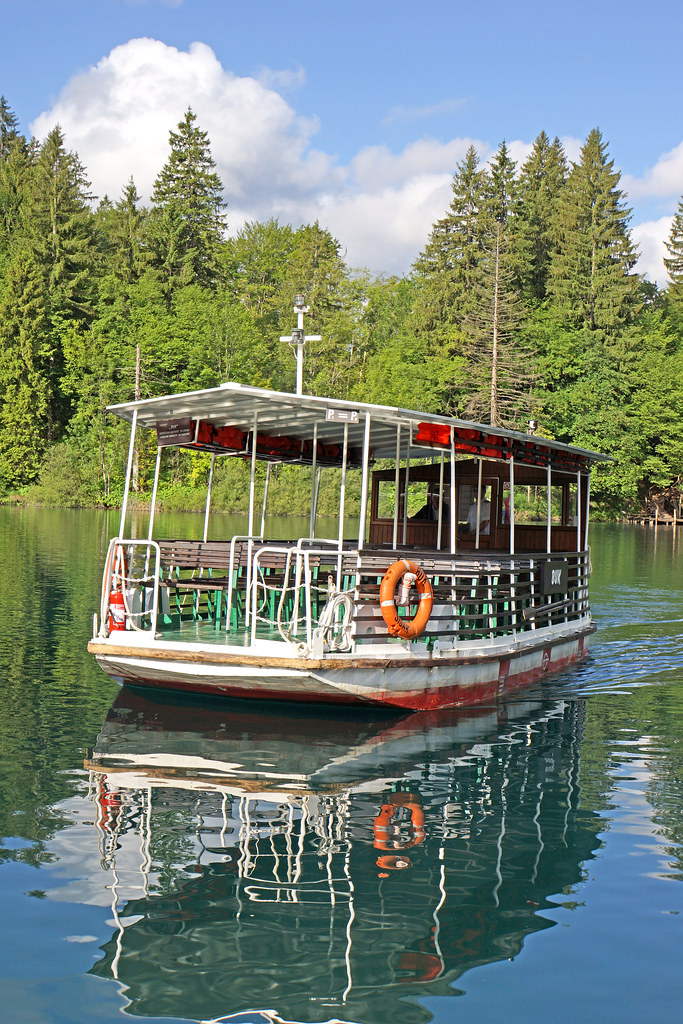 Covered passenger boat on a glassy lake, framed by a dense pine forest and its mirror-like reflections