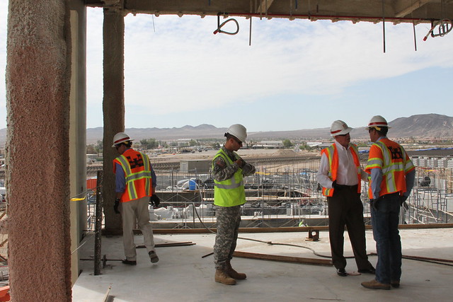 Four people in hard hats and high-visibility vests stand on a concrete deck at Fort Irwin with scaffolding and desert hills in the background during a Weed Army Hospital project briefing.