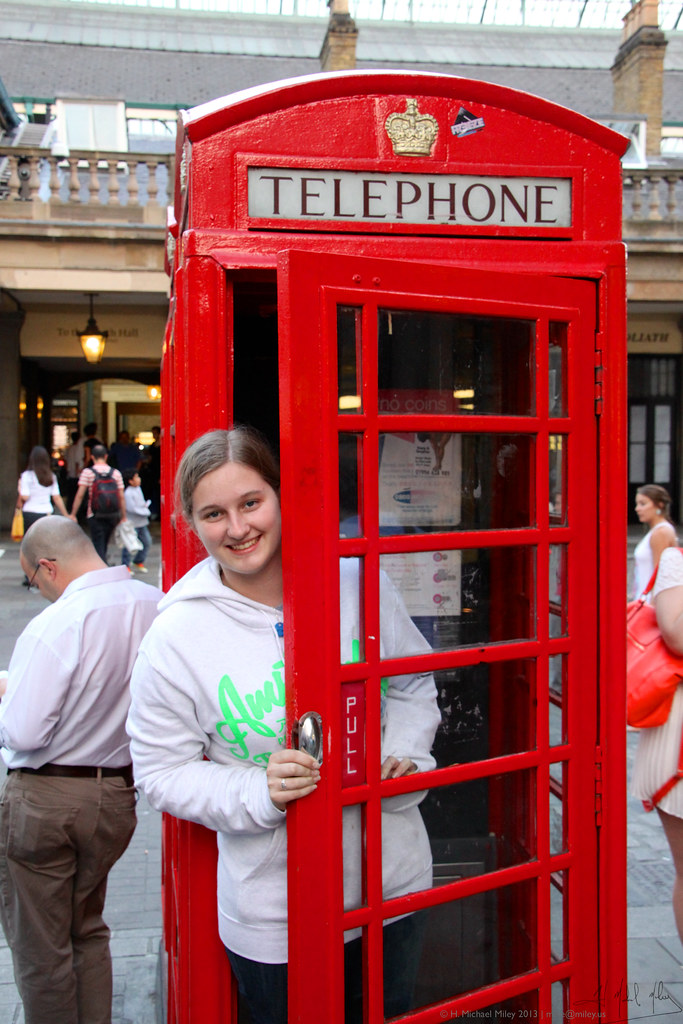 Portrait of a girl in a light gray hoodie leaning from a bright red British telephone booth amid a busy city street