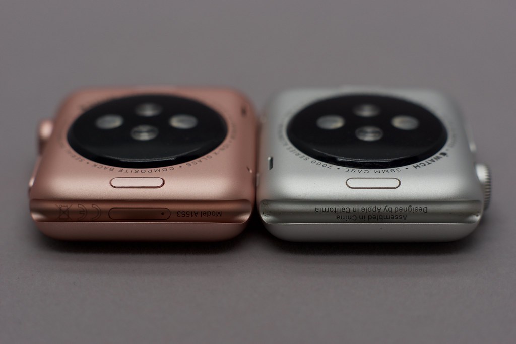 Close-up of Rose Gold and Silver Apple Watch backs on a gray surface, highlighting the circular sensor array and precision machining