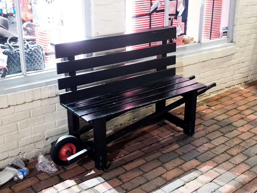 Wheelbarrow-Style Black Wooden Bench on a Brick Sidewalk by a Storefront Window