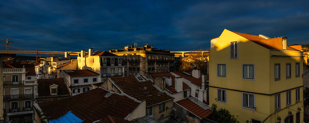 Golden-hour panorama from a Lisbon rooftop patio toward a sunlit yellow façade, with diagonal rooflines and red-tiled rooftops