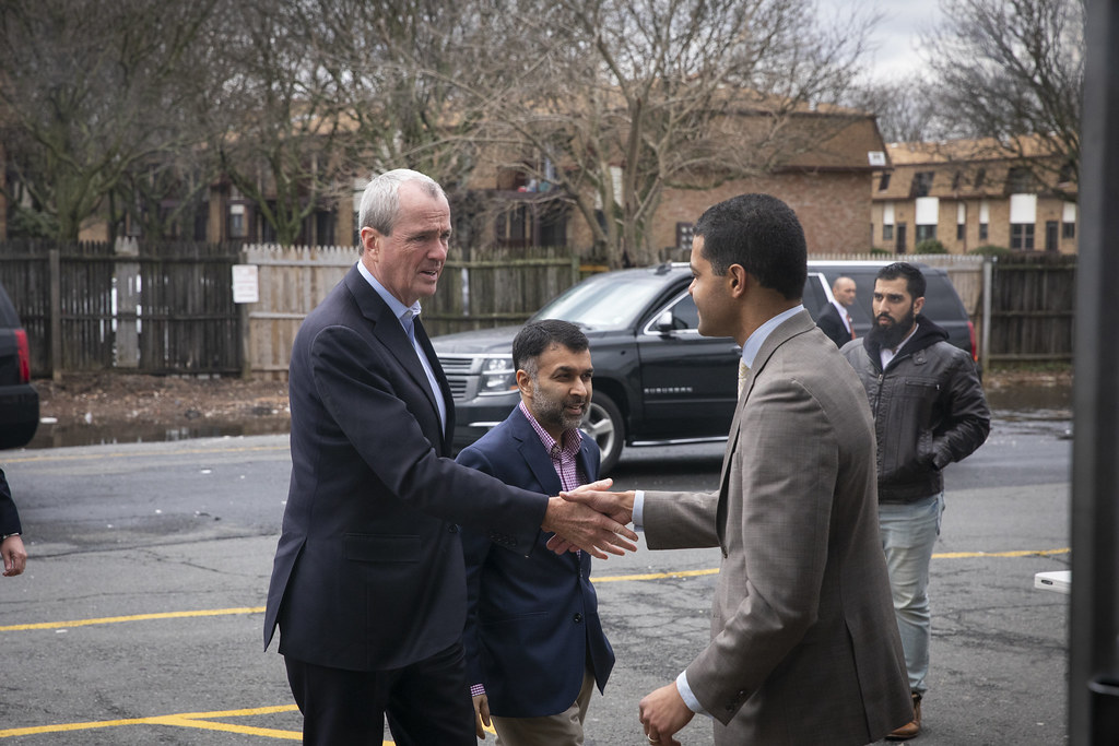 Two suited men exchange a handshake on a city street, with bystanders and parked vehicles in a neighborhood backdrop
