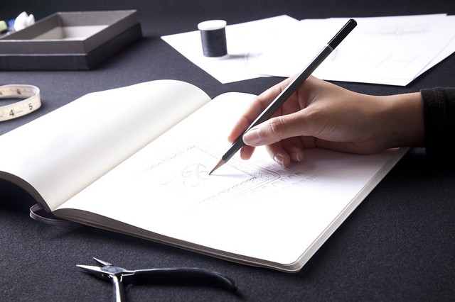 Hand sketching a decorative design in an open notebook on a dark drafting desk with measuring tape and pliers nearby.