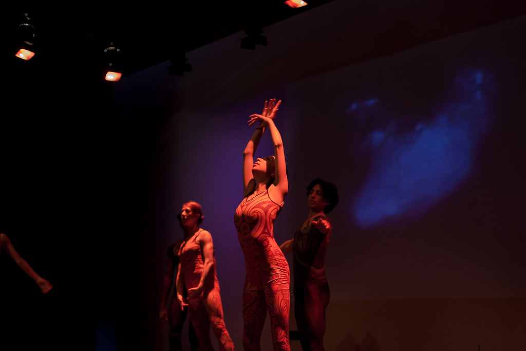 Vertical Silhouette Under Violet-Blue Projection: Lead Dancer in a Red Patterned Unitard Extends Arms Overhead, Backed by Two Dancers Under Crimson Lighting