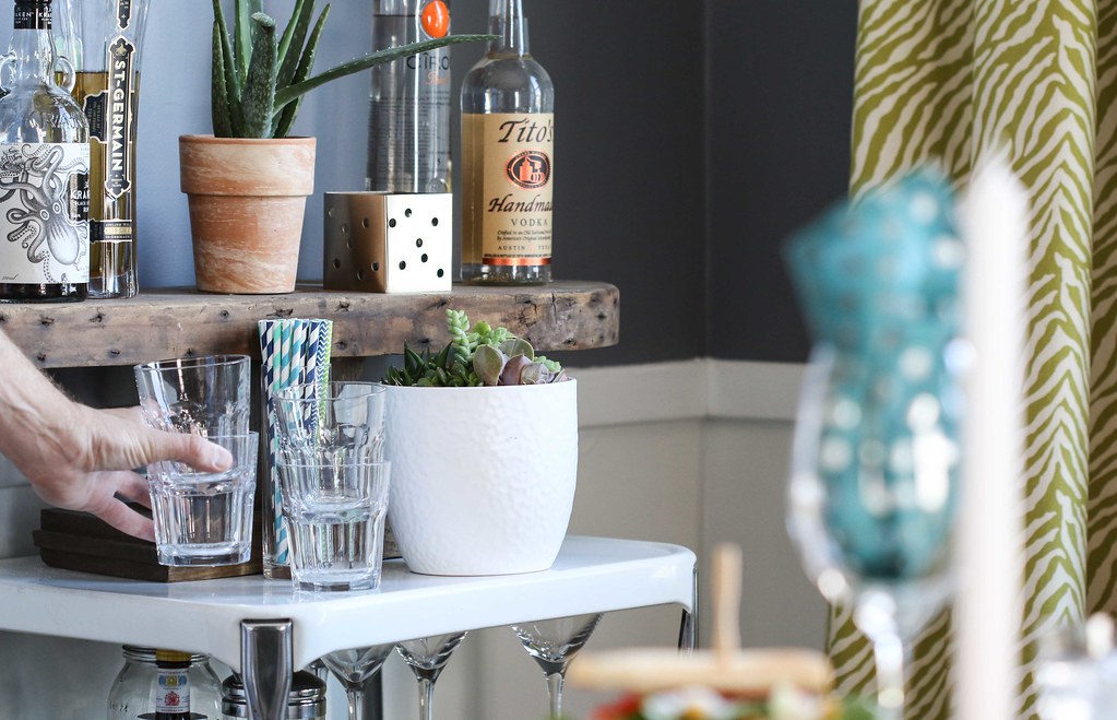 Succulents in a white textured pot on a rustic bar cart beside glassware, striped straw cups, and liquor bottles