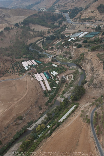 Aerial view of a dry, hilly valley with a winding river, terraced fields, and long poly-tunnel greenhouses along the riverbank.