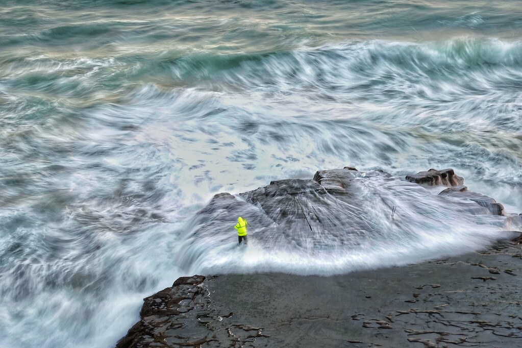 Tempting Fate on Flat Rock: A lone figure braves surging seas along a jagged shoreline